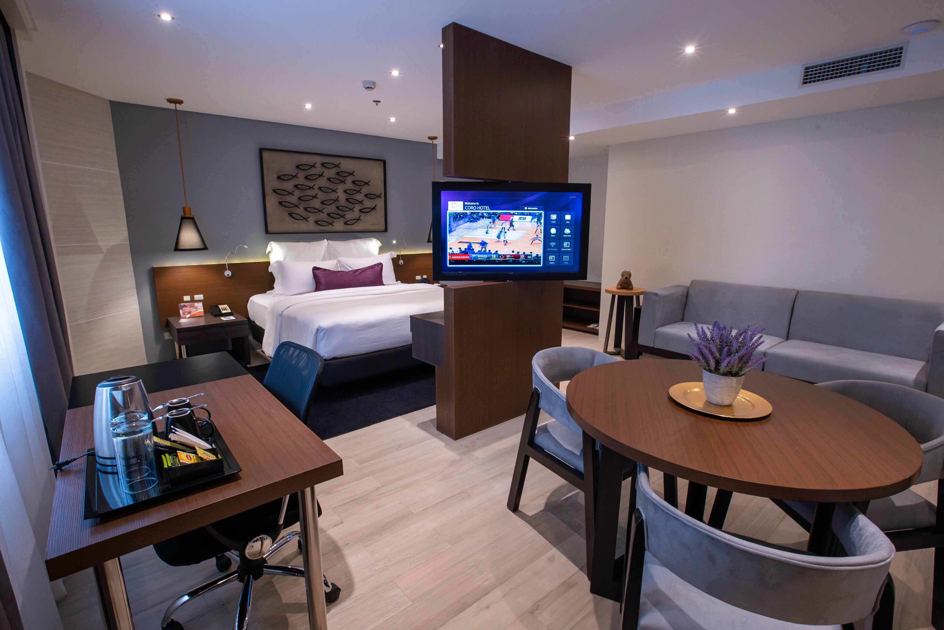 Quarto de hotel moderno com uma cama, uma TV fixada na parede, área de estar com uma mesa e cadeiras, e uma pequena mesa lateral com uma planta.