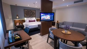Quarto de hotel moderno com uma cama, uma TV fixada na parede, área de estar com uma mesa e cadeiras, e uma pequena mesa lateral com uma planta.