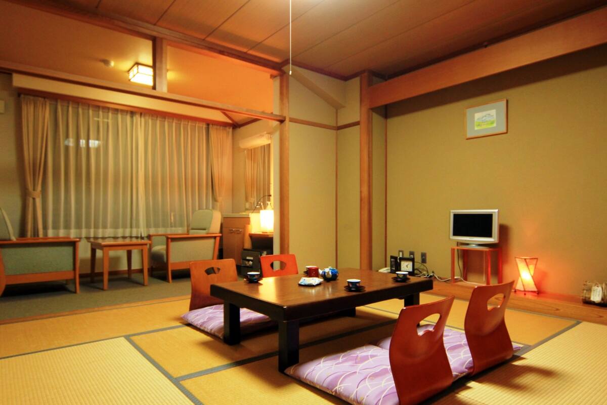 Um quarto em estilo japonês tradicional, com tatames, uma mesa baixa com almofadas e uma televisão em um móvel.