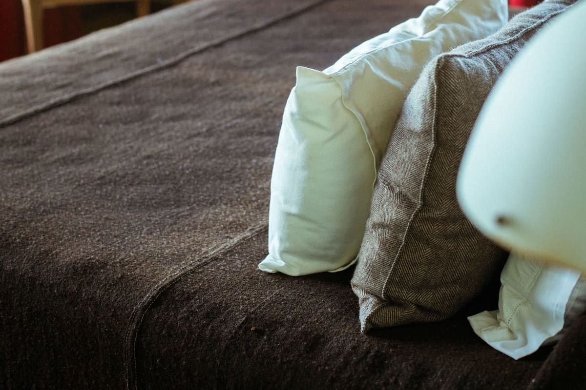 Cama bem arrumada com manta marrom, almofadas brancas e cabeceira. Ao fundo, cômoda de madeira com gavetas e uma cadeira.