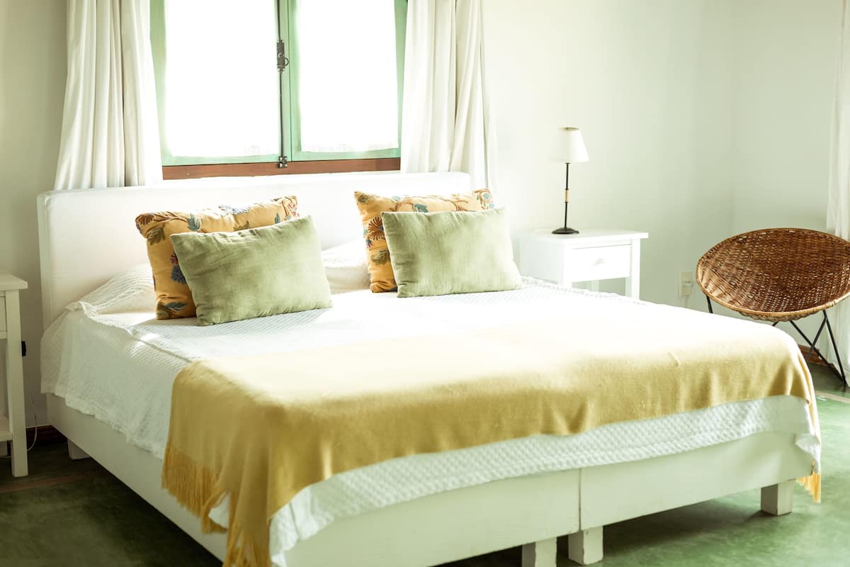 Uma cama bem arrumada com cabeceira branca, duas almofadas e um cobertor amarelo. Há uma cadeira de vime e uma mesa de cabeceira pequena com uma luminária.