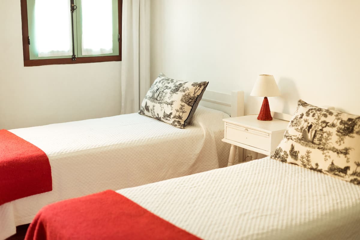 Duas camas de solteiro com roupa de cama vermelha e branca, um criado-mudo com uma luminária e uma janela com cortinas.