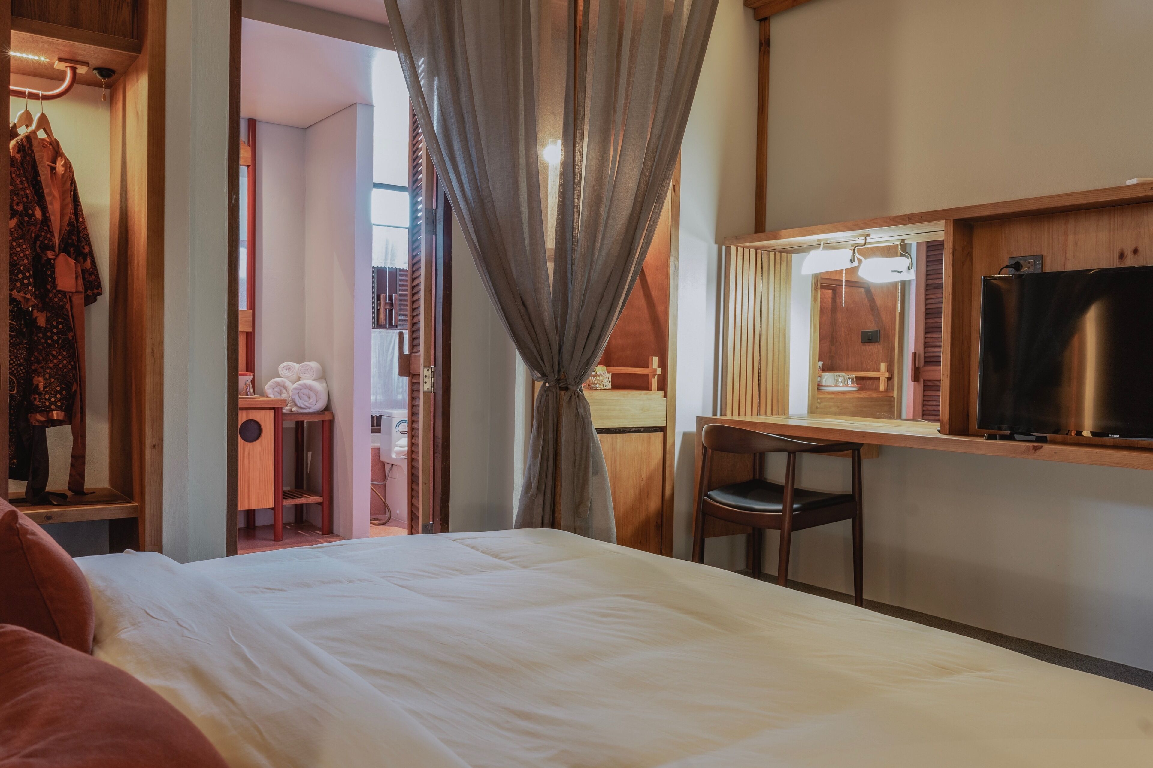 Quarto de hotel com cama, escrivaninha com cadeira, televisão e guarda-roupa.