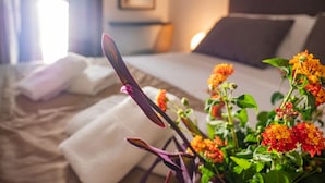 Uma cama bem arrumada com lençóis brancos, uma mesa de cabeceira com abajur, e um arranjo de flores sobre a mesa.