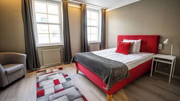 Quarto com cama vermelha, tapete cinza e vermelho, poltrona cinza e mesa de cabeceira branca com abajur.