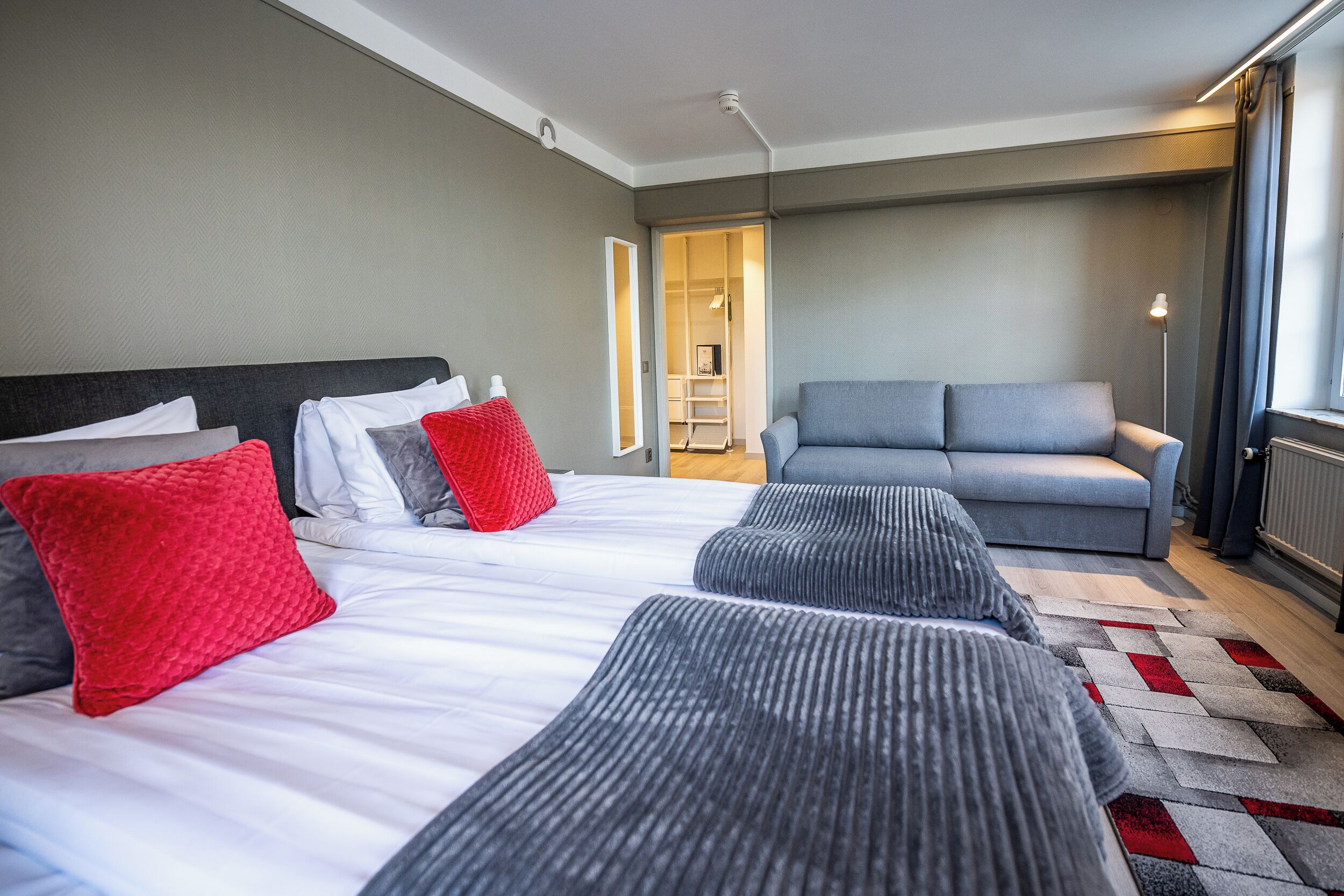 Quarto de hotel com cama, sofá cinzento e vista para um corredor.