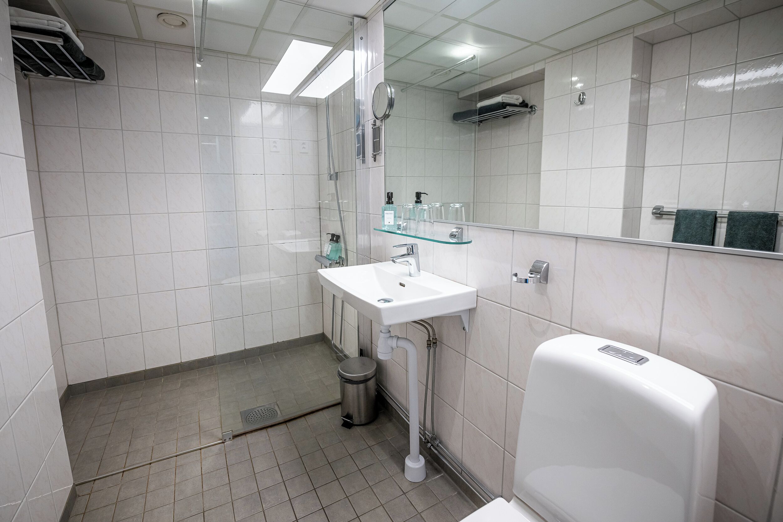 Uma casa de banho moderna com uma sanita branca, prateleiras de vidro, um espelho e uma área de duche.
