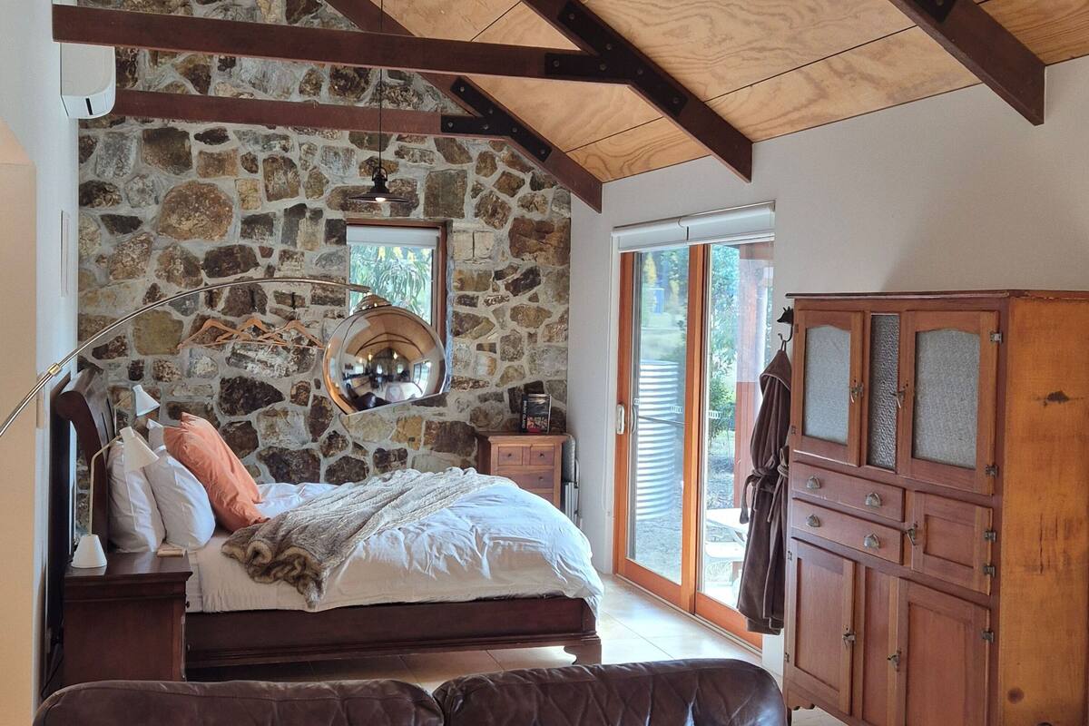 Quarto com parede de pedra, vigas de madeira, cama, cômoda e sofá de couro.