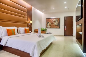 Quarto de hotel com cama grande, cabeceira de madeira, um quadro na parede, uma planta e uma porta.