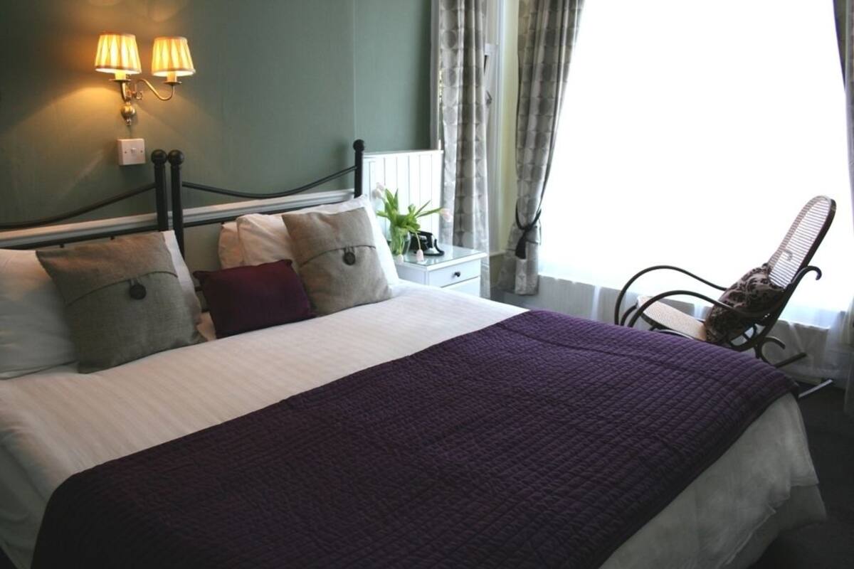 Uma cama bem arrumada com um cobertor roxo, uma mesa de cabeceira com abajur e uma cadeira perto da janela.