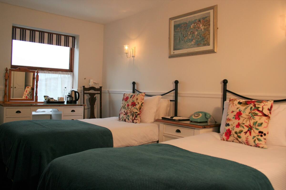 Quarto com duas camas de solteiro, mesa de cabeceira com telefone, cômoda, espelho e janela com cortinas.