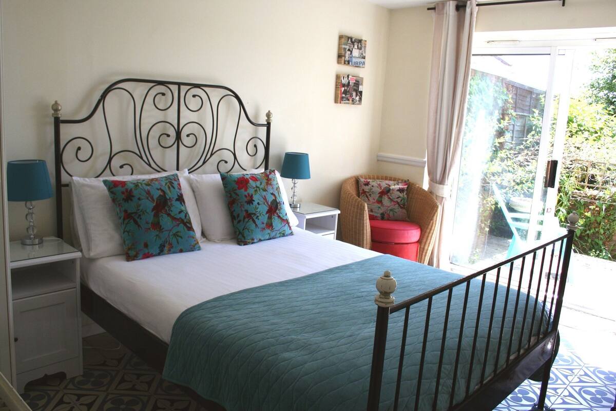 Cama bem arrumada com colcha verde-água e almofadas florais, uma pequena mesa de cabeceira com abajur, e vista para o verde através de uma porta de correr de vidro.