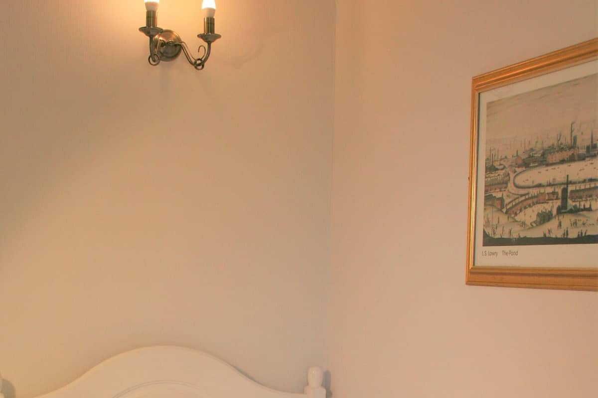 Cama bem arrumada com lençóis brancos e travesseiro verde-água, luminária fixada na parede e quadro emoldurado.