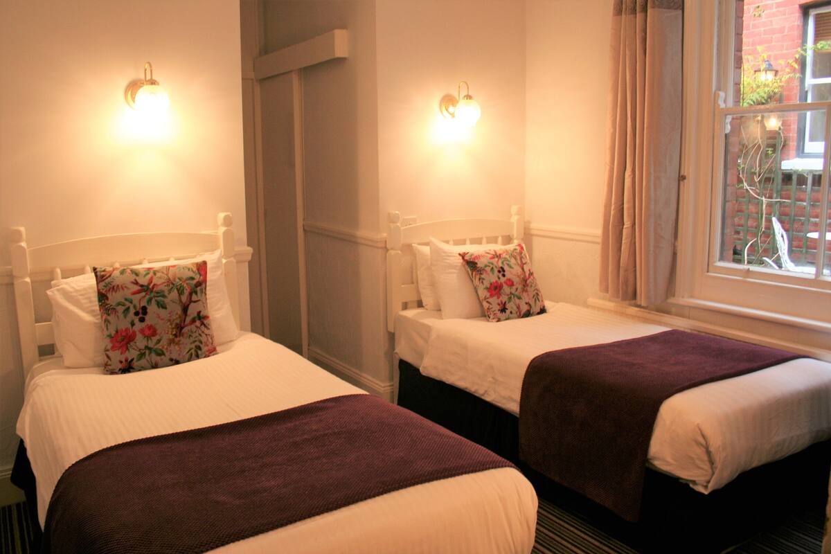 Duas camas de solteiro com cabeceiras brancas e almofadas estampadas com flores, uma janela com vista para o exterior e luminárias fixadas na parede.