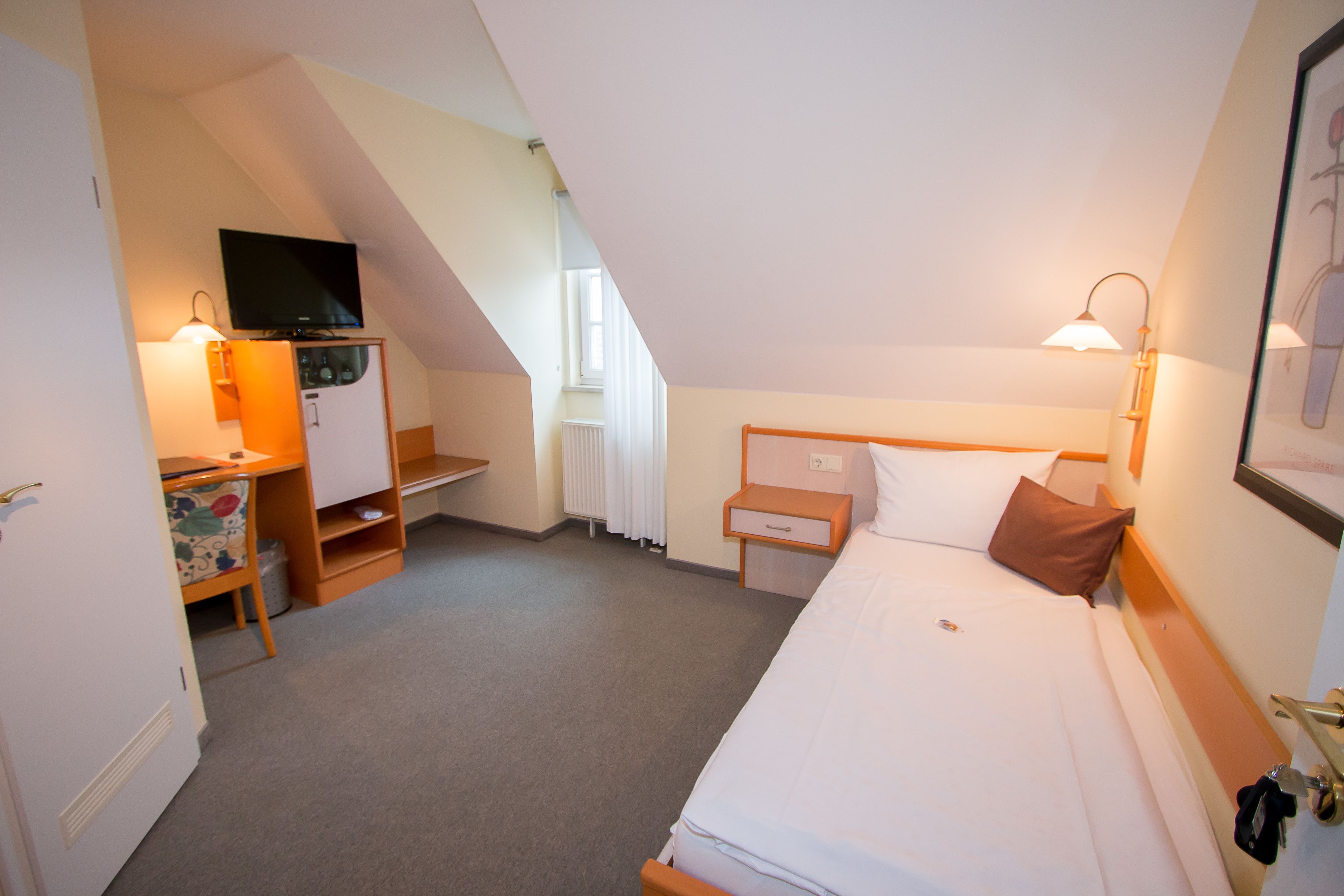 Quarto de hotel com cama, secretária e cadeira, televisão e janela.