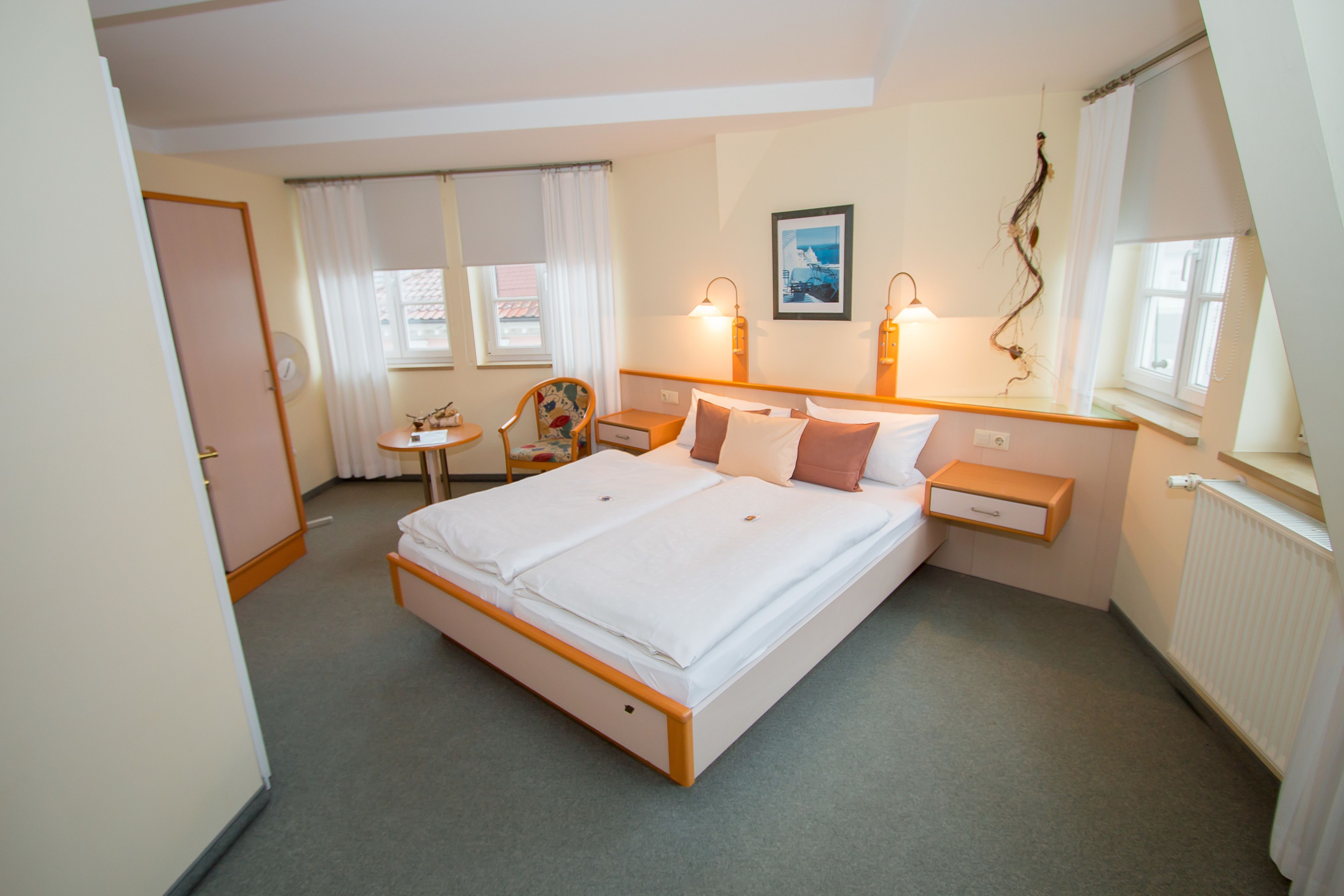 Quarto de hotel com uma cama de solteiro, mesas de cabeceira, uma cadeira, uma pequena mesa e uma janela com cortinas.