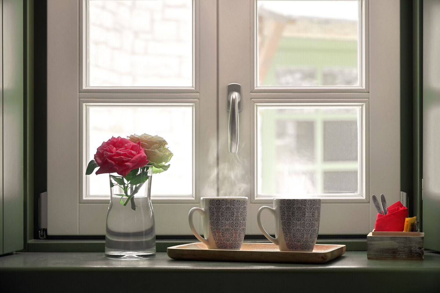 Uma janela com um vaso de flores, duas chávenas estampadas e um tabuleiro de madeira.