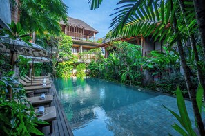 Uma área de piscina com espreguiçadeiras e um deck de madeira, cercada por vegetação exuberante e um edifício com varanda.