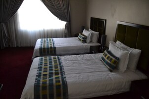 Duas camas de solteiro com cabeceiras, uma janela com cortinas e um tapete vermelho.