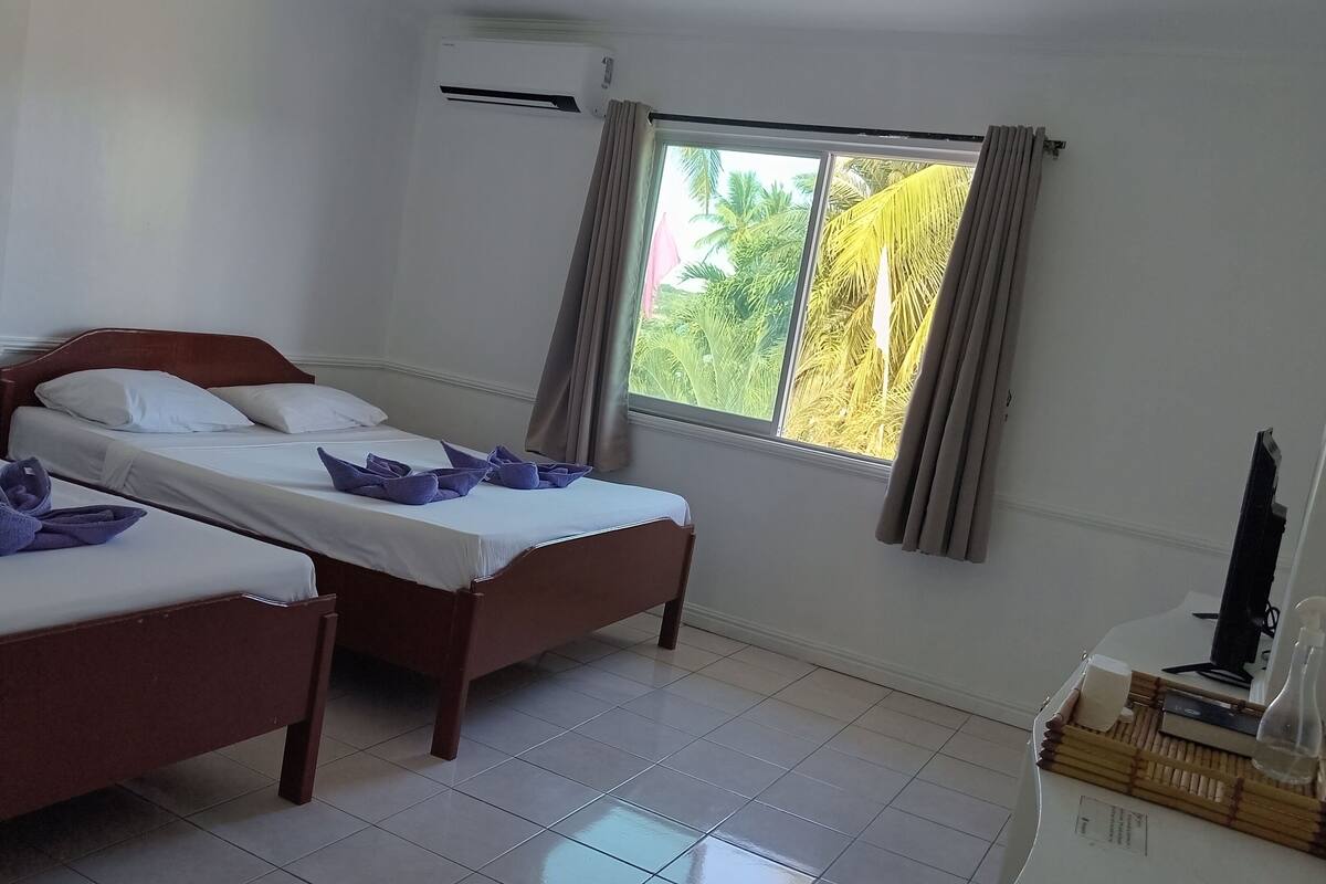 Quarto de hotel com duas camas, janela com cortinas, televisão e uma mesinha de apoio com produtos de higiene pessoal.
