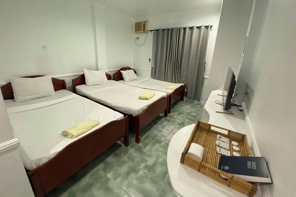 Quarto de hotel com três camas simples, uma pequena mesa com um computador e um ar condicionado fixado na parede.