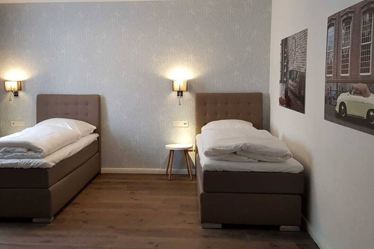 Quarto de hotel com duas camas de solteiro, uma pequena mesa de cabeceira, uma luminária fixada na parede e um quadro emoldurado.