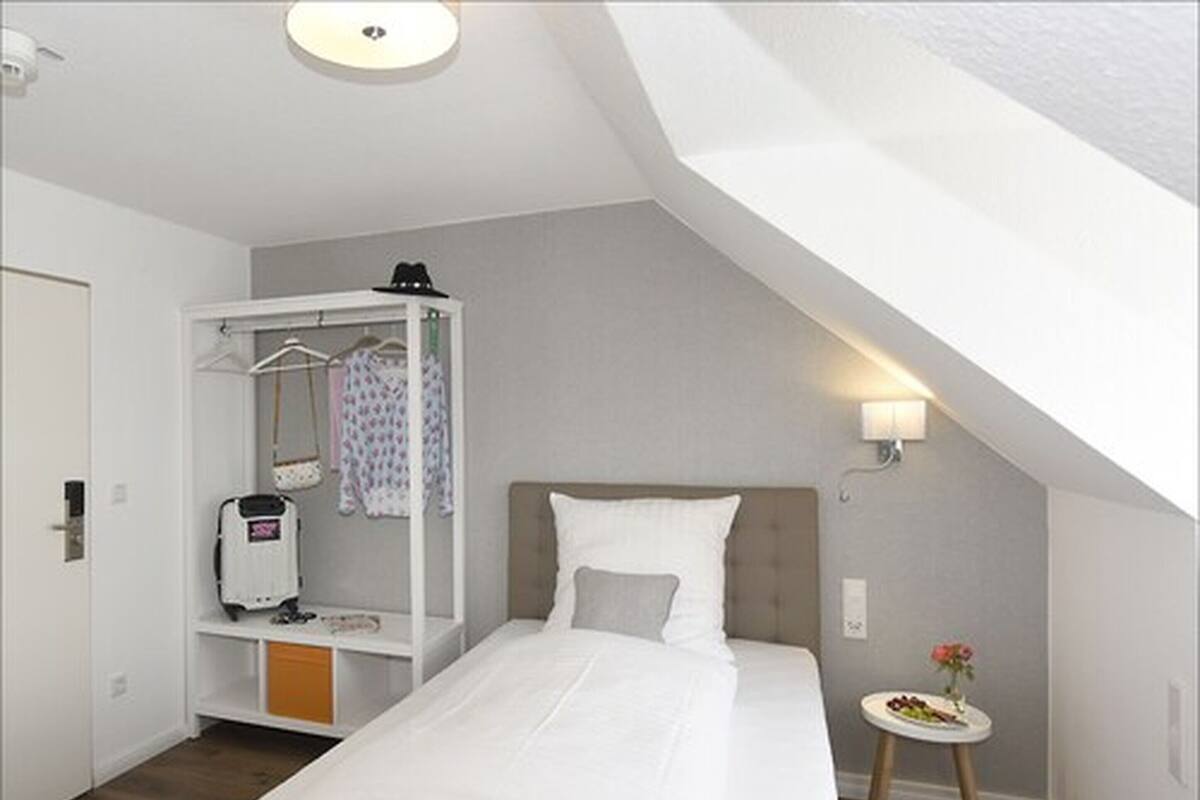 Quarto com cama, guarda-roupa, uma pequena mesa e um abajur fixado na parede.