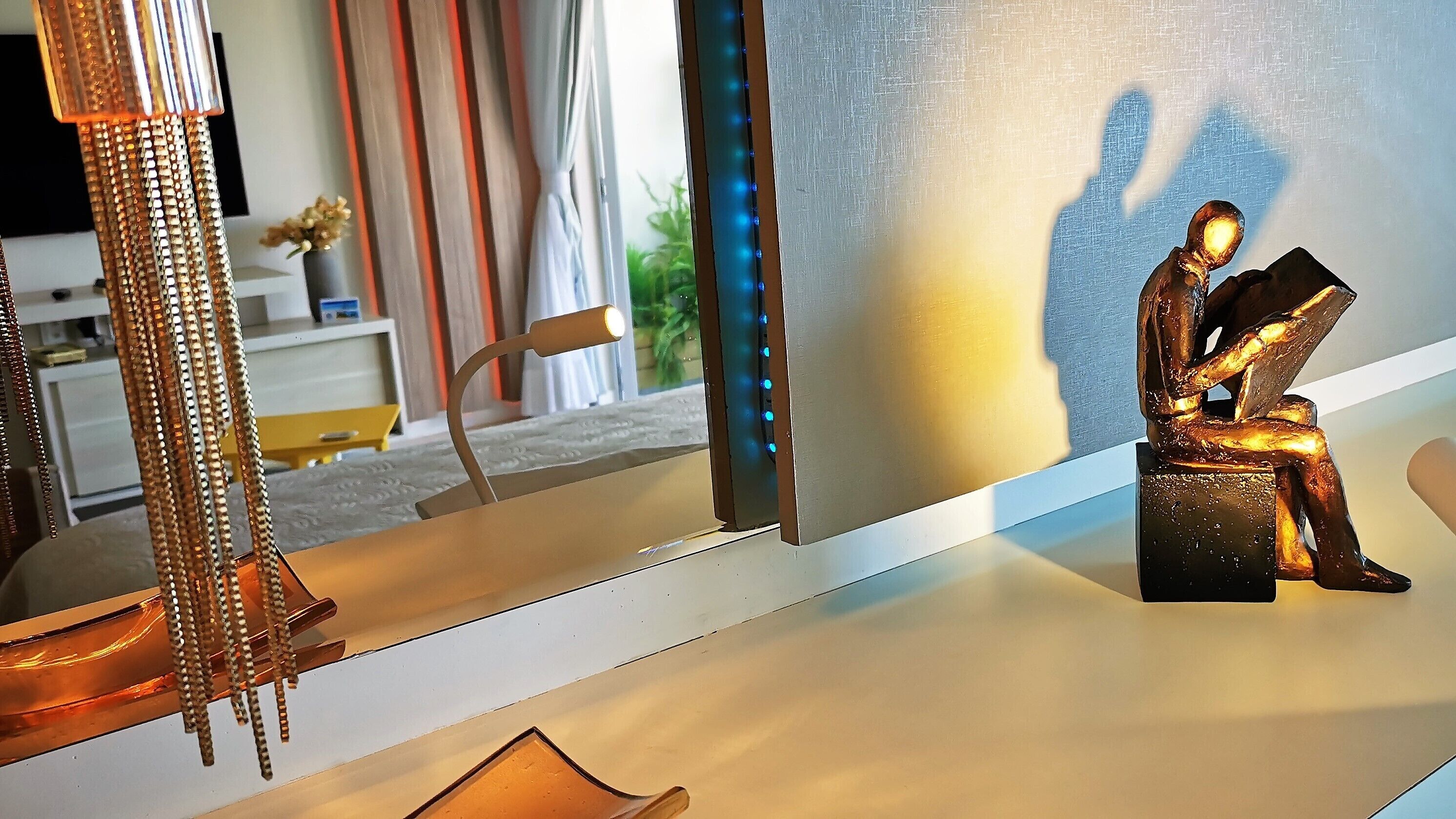 Quarto com uma escultura metálica decorativa, uma mesa pequena com um vaso e uma parede com a sombra de uma pessoa.