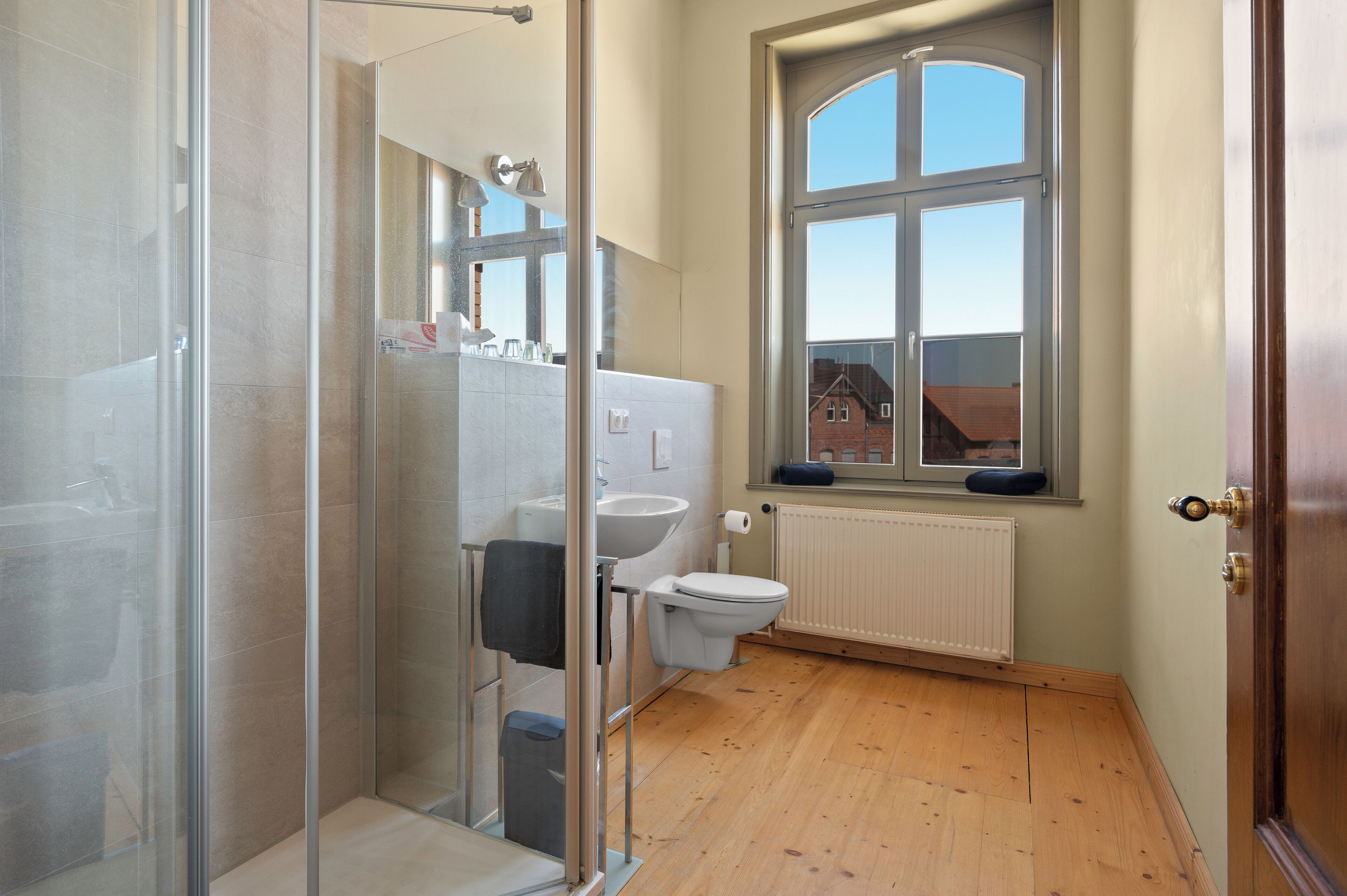Um banheiro moderno com box de vidro, pia branca e vaso sanitário. Há uma janela com vista para um edifício.