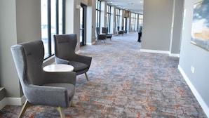 Um saguão de hotel com poltronas cinzas, piso carpete e grandes janelas, com um quadro na parede.
