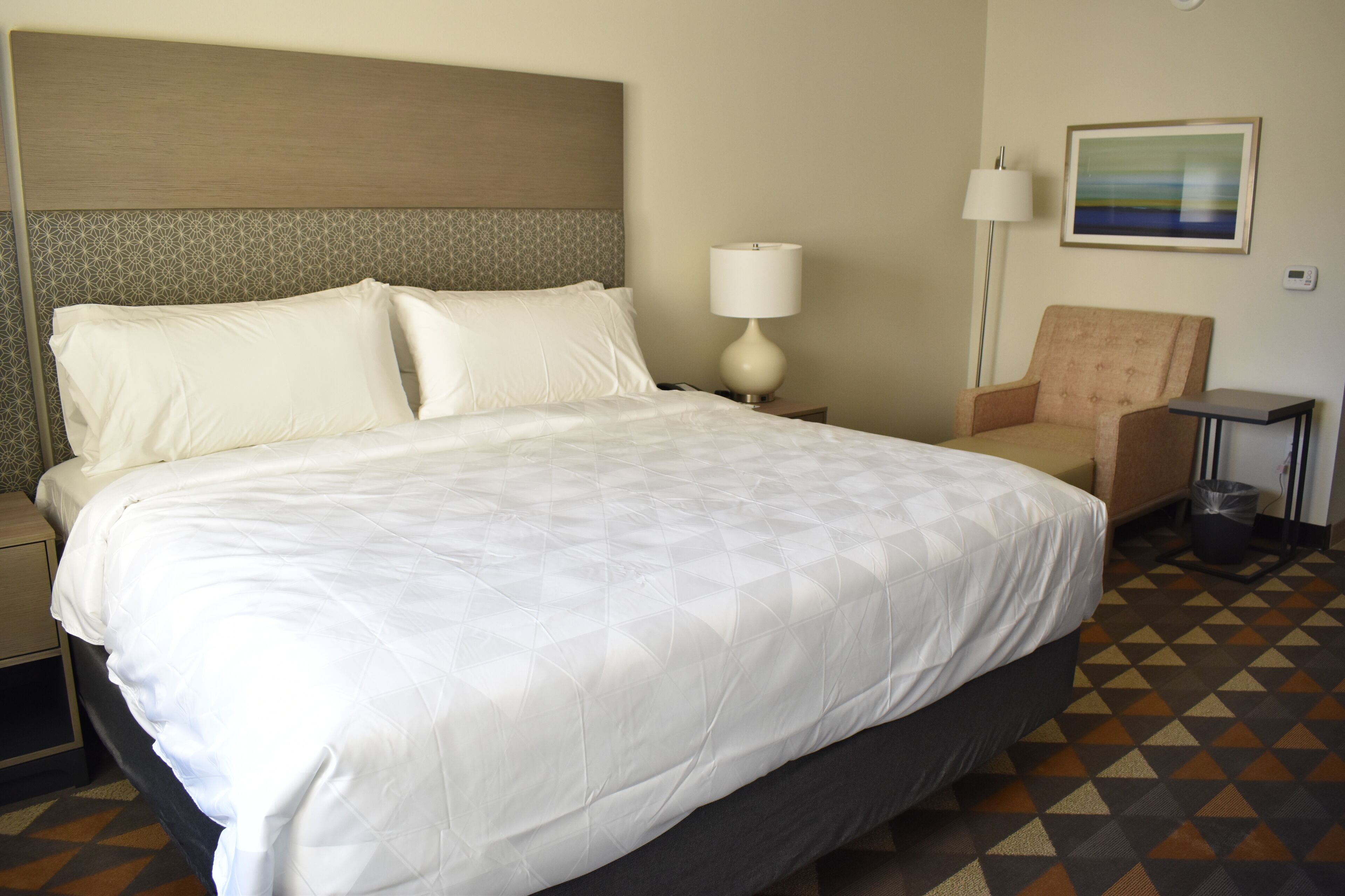 Quarto de hotel com cama grande, mesa de cabeceira, cadeira, luminária e um quadro na parede.