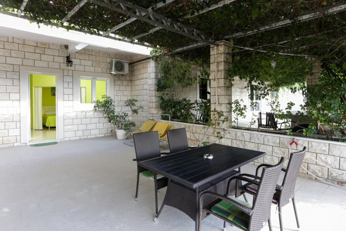 Um pátio externo com mesa e cadeiras, cercado por paredes de pedra e vegetação.