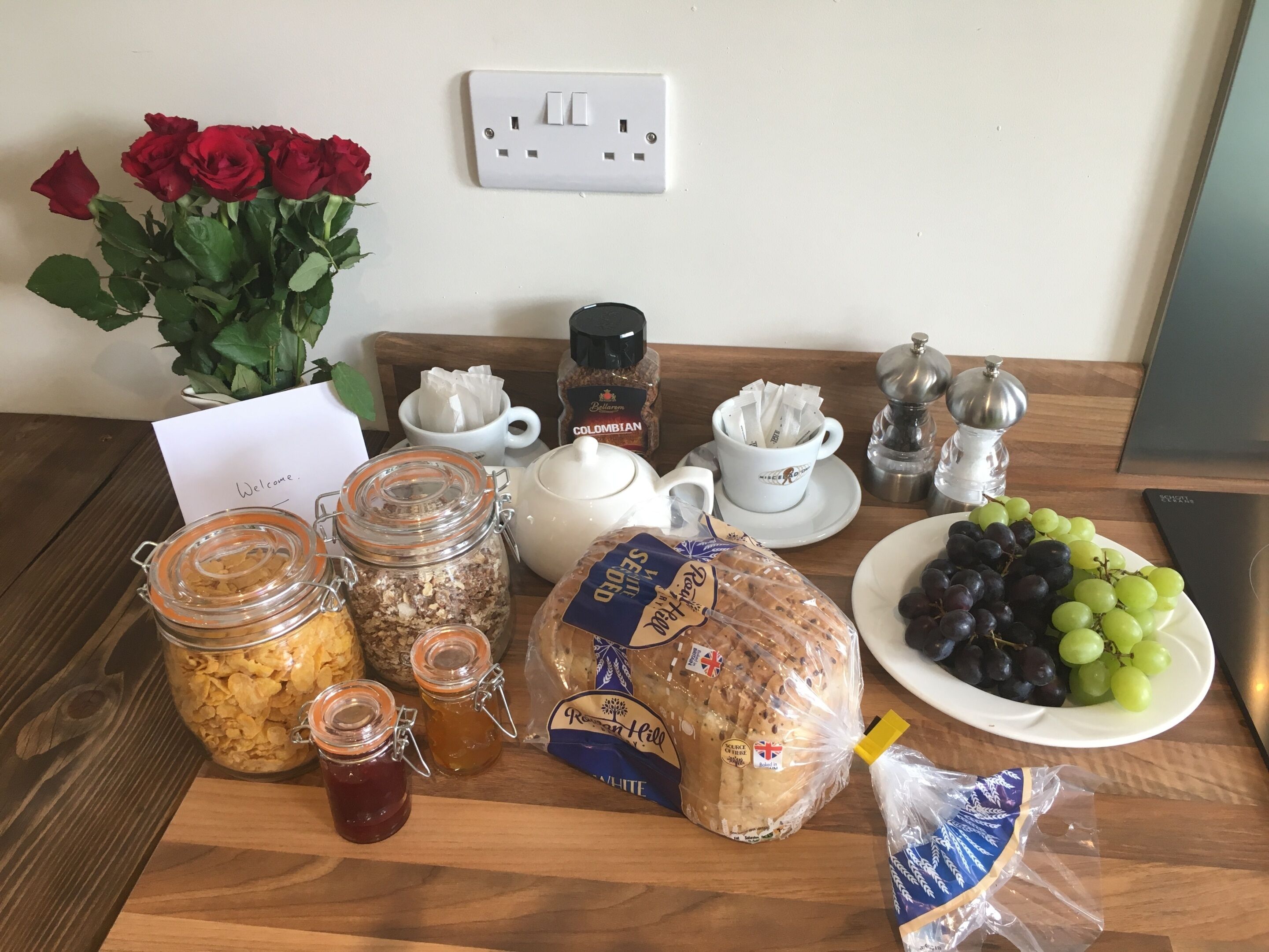 Uma mesa de café da manhã com diversos alimentos, uma cafeteira, um açucareiro e um prato de uvas.