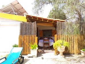 Uma pérgola de madeira com mesa e cadeiras por baixo, rodeada por plantas em vasos e uma cadeira azul.