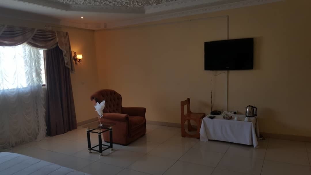 Quarto de hotel com uma poltrona marrom, uma pequena mesa com toalha branca, uma televisão de tela plana na parede e uma janela com cortinas.