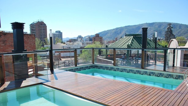 Piscina ao ar livre com deck de madeira, rodeada por guarda-corpo de vidro, com vista para uma paisagem urbana e montanhas.