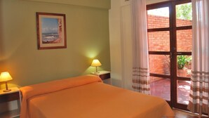Quarto com cama, mesa de cabeceira com luminária, quadro pendurado na parede e vista através de uma porta de vidro para área externa com parede de tijolos e plantas.