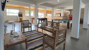 Ampla sala de estar com um sofá, mesa de centro e tapete listrado. Cozinha visível ao fundo, com balcão e extintor de incêndio.