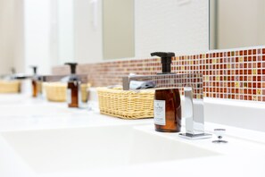 Casa de banho com várias lavatórios, cada um com dispensador de sabonete e um copo. Há uma cesta de vime colocada sobre um dos lavatórios.