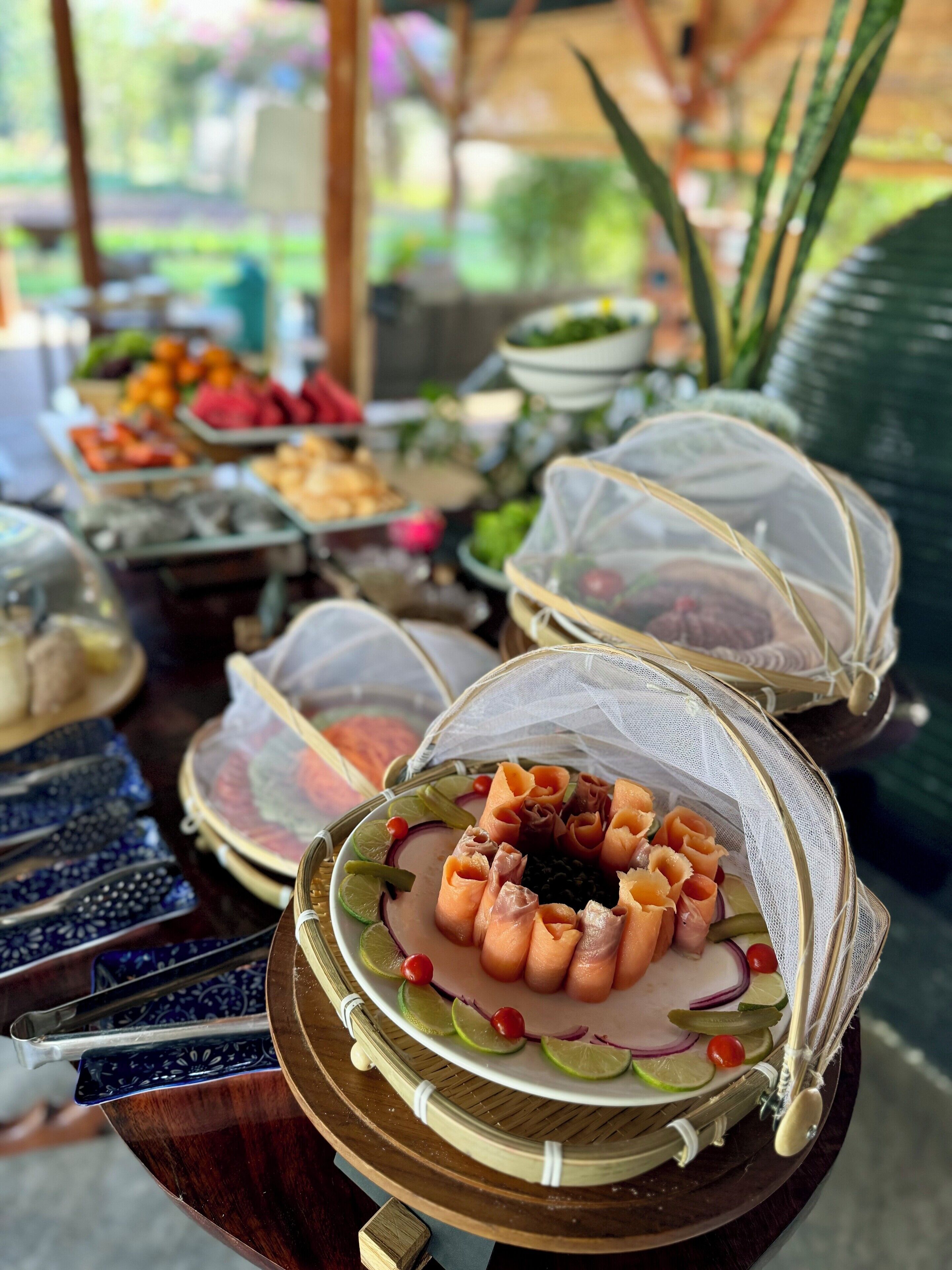 Mesa de buffet com diversos alimentos, incluindo um prato central com carne e acompanhamentos, cercado por outros pratos e recipientes tampados.