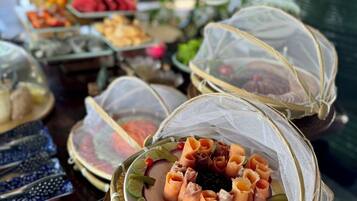 Mesa de buffet com diversos alimentos, incluindo um prato central com carne e acompanhamentos, cercado por outros pratos e recipientes tampados.