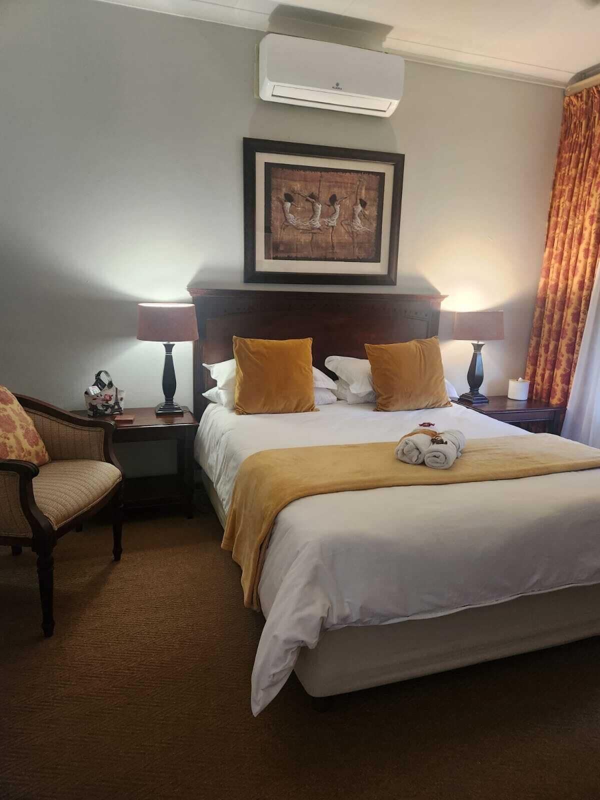 Quarto de hotel com uma cama grande, duas mesas de cabeceira com luminárias, uma cadeira, ar condicionado e uma obra de arte emoldurada na parede.