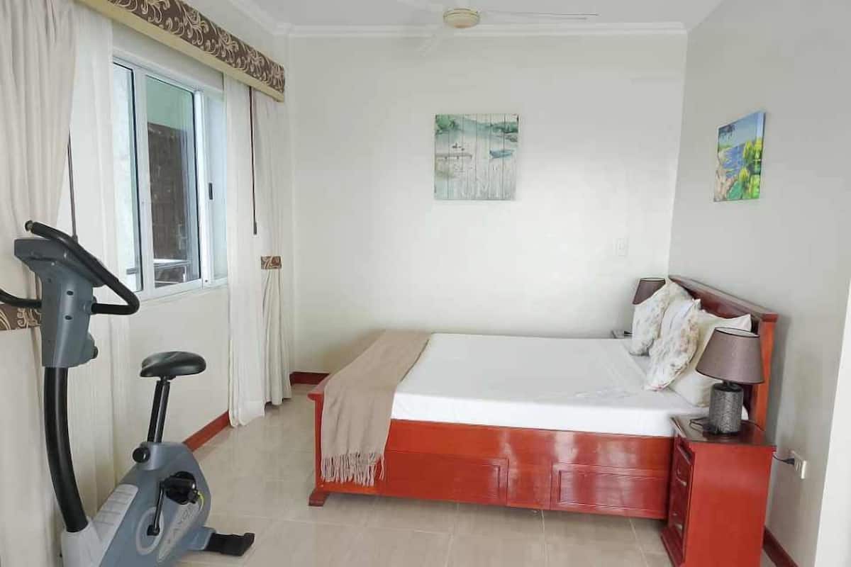 Quarto com cama, mesa de cabeceira, luminária, quadro na parede e janela com cortinas.