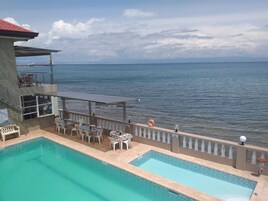 Área da piscina com vista para o mar, mobiliário de exterior e um edifício com telhado vermelho.
