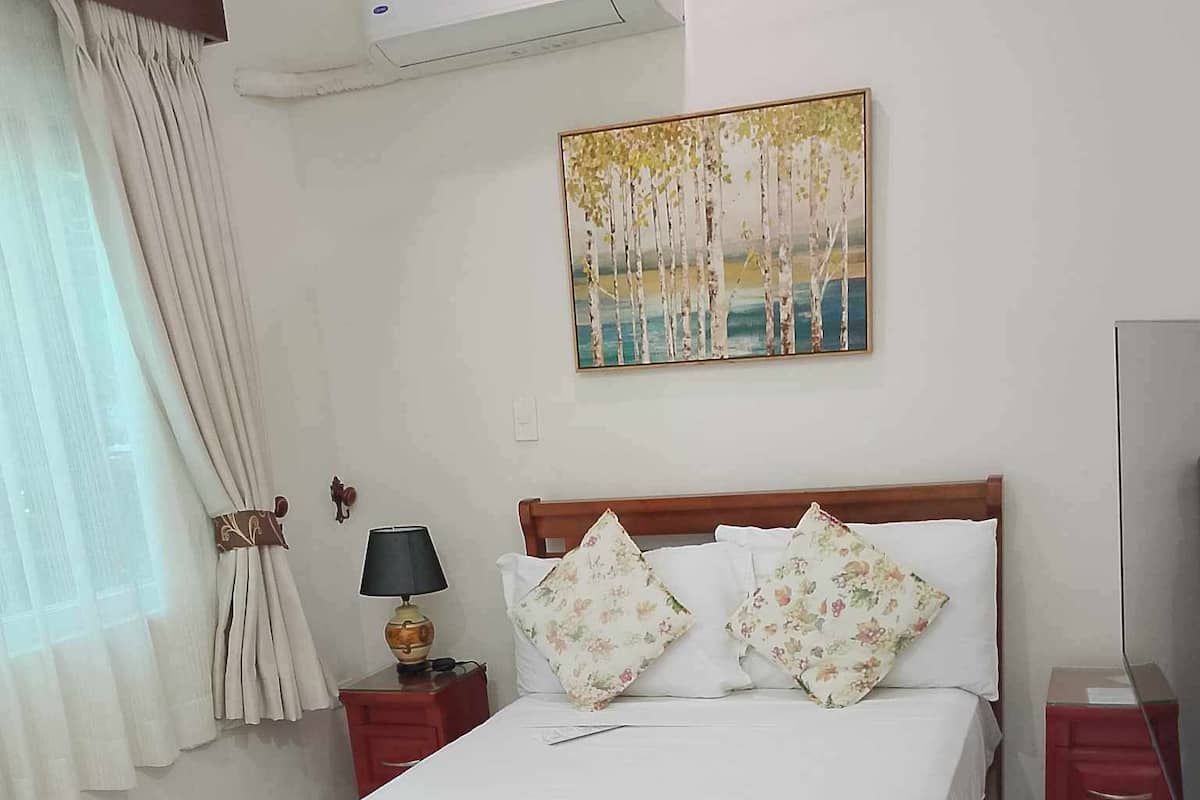 Uma cama bem arrumada com estrutura de madeira, uma mesa de cabeceira com abajur, e um quadro acima da cama.