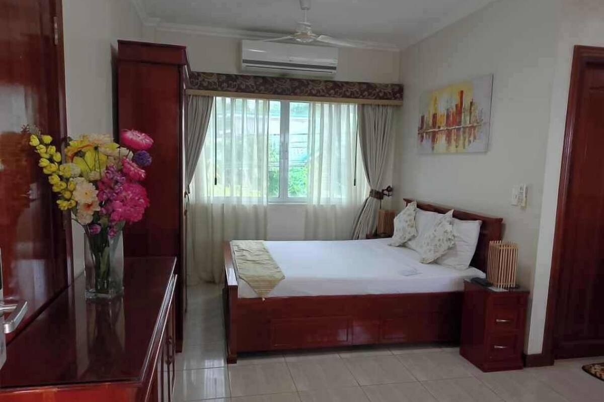 Quarto de hotel com cama, cómoda de madeira, um vaso de flores e janela com cortinas.