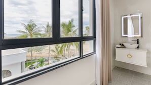 Casa de banho moderna com uma janela grande, toucador branco e vista para uma praia e palmeiras.