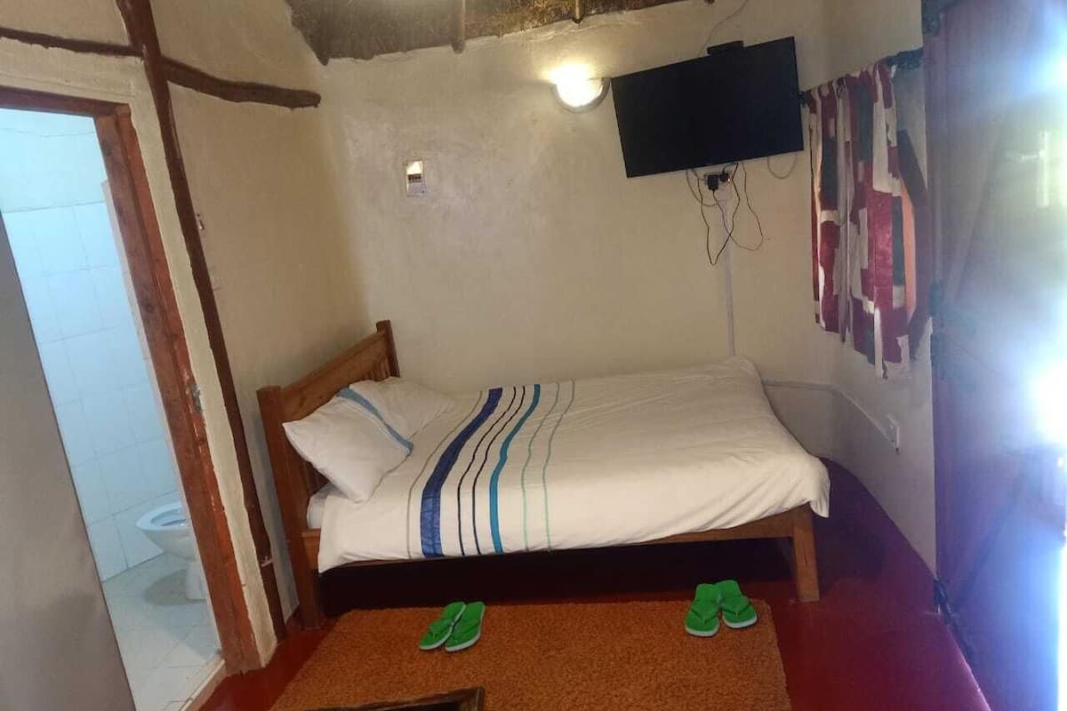 Quarto pequeno com uma cama de solteiro, cabeceira de madeira, colcha branca e uma TV fixada na parede.