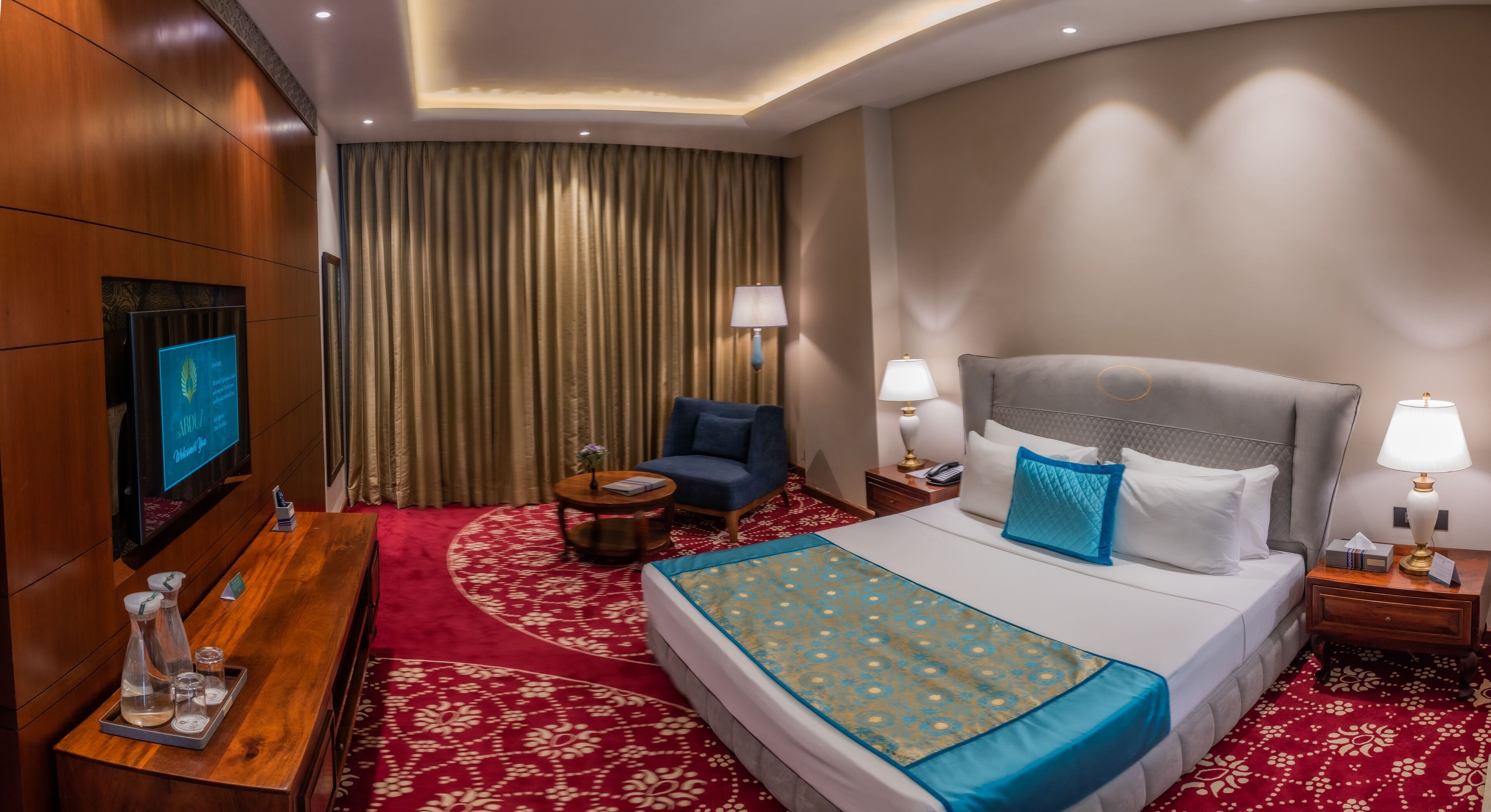 Quarto de hotel com uma cama grande, televisão na parede, escrivaninha com cafeteira e um tapete estampado em vermelho.