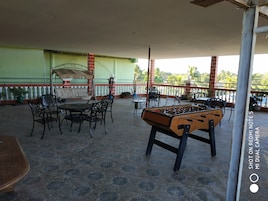 Uma área externa com mesas, cadeiras, uma mesa de futebol e vista para o verde.
